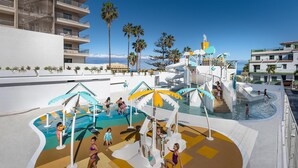 Área de parque aquático com toboáguas e piscinas, cercada por palmeiras e edifícios.