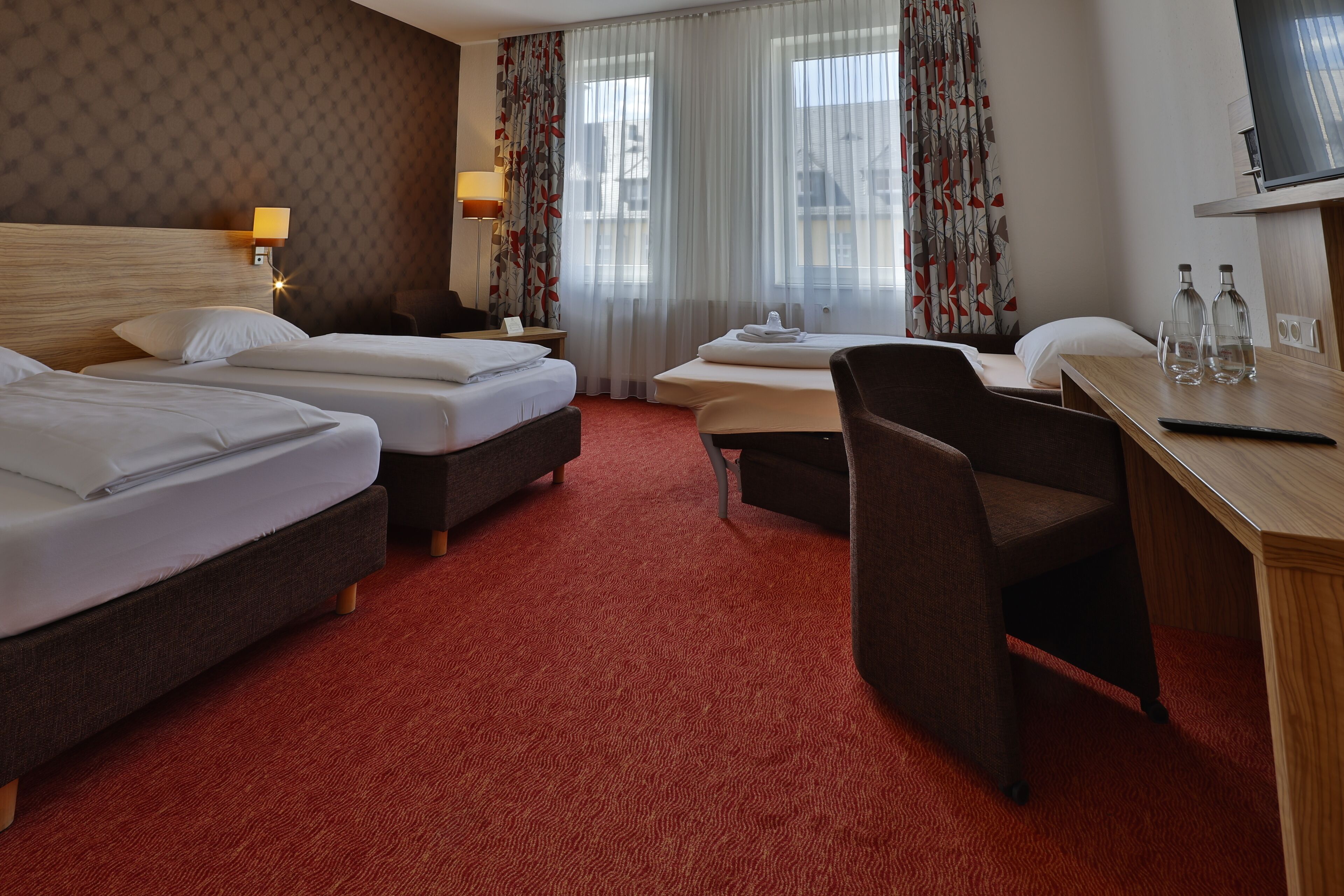 Quarto de hotel com duas camas, uma escrivaninha, uma cadeira, uma televisão e uma janela com cortinas.