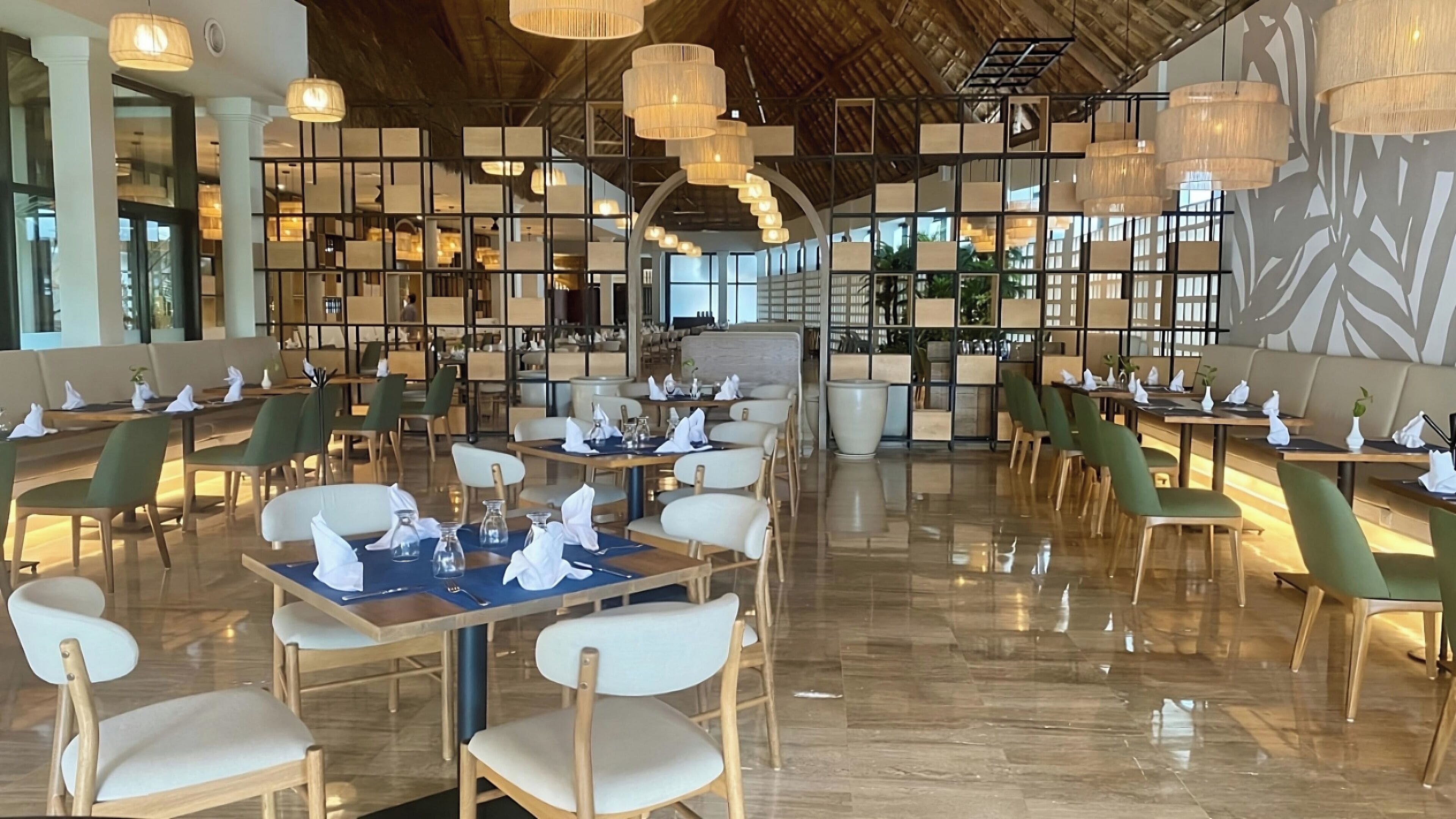 Um restaurante amplo com tetos altos, lustres modernos e piso limpo e bem cuidado. Mesas organizadas com guardanapos brancos e copos.