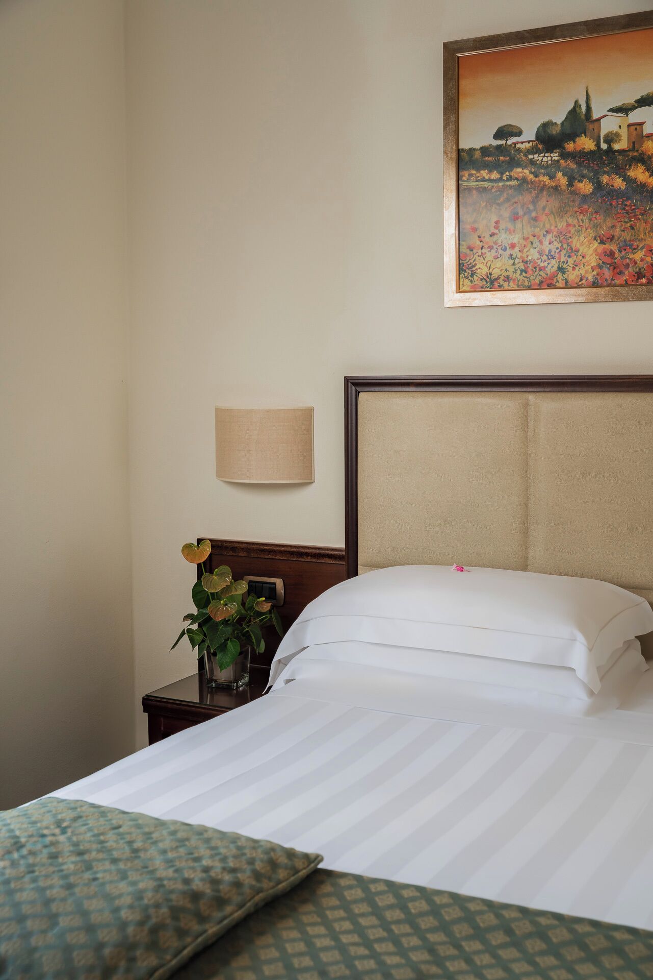 Quarto de hotel com cama, mesa de cabeceira com abajur, um quadro na parede e uma planta.