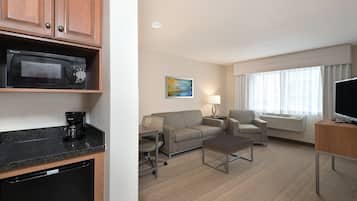 Quarto de hotel com cozinha compacta, sofá, mesa de centro, televisão e janela com cortinas.