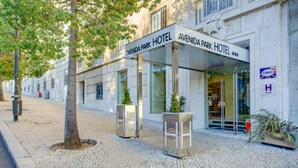 Exterior do Hotel Avenida Park, com calçada de pedra, grandes vasos de plantas e vista clara da entrada.