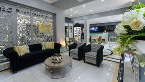 Um hall de hotel moderno com um sofá preto, poltronas estampadas, uma mesa de centro e uma parede decorativa.