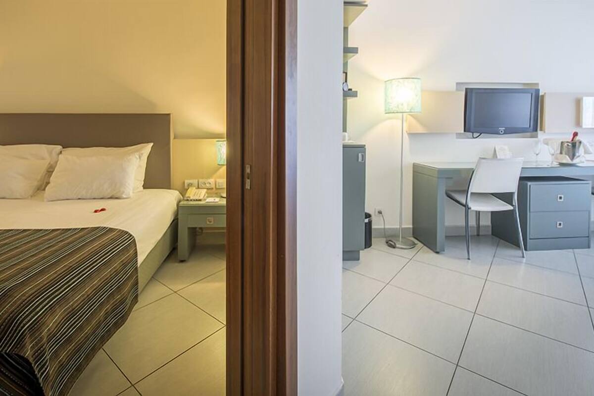 Quarto de hotel com cama, mesas de cabeceira, escrivaninha com cadeira, televisão e abajur.