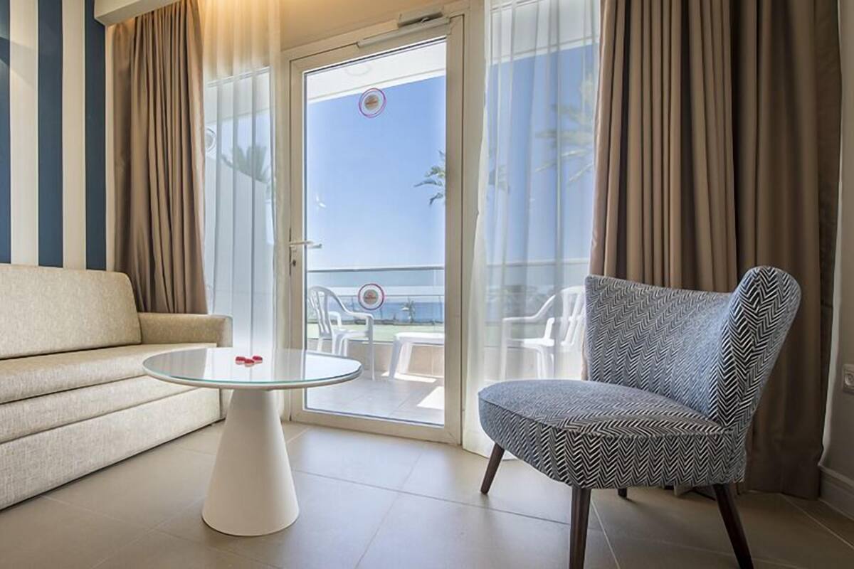 Quarto com sofá, poltrona estampada, mesa de centro de vidro e porta de correr que dá para área externa com piscina e cadeiras.