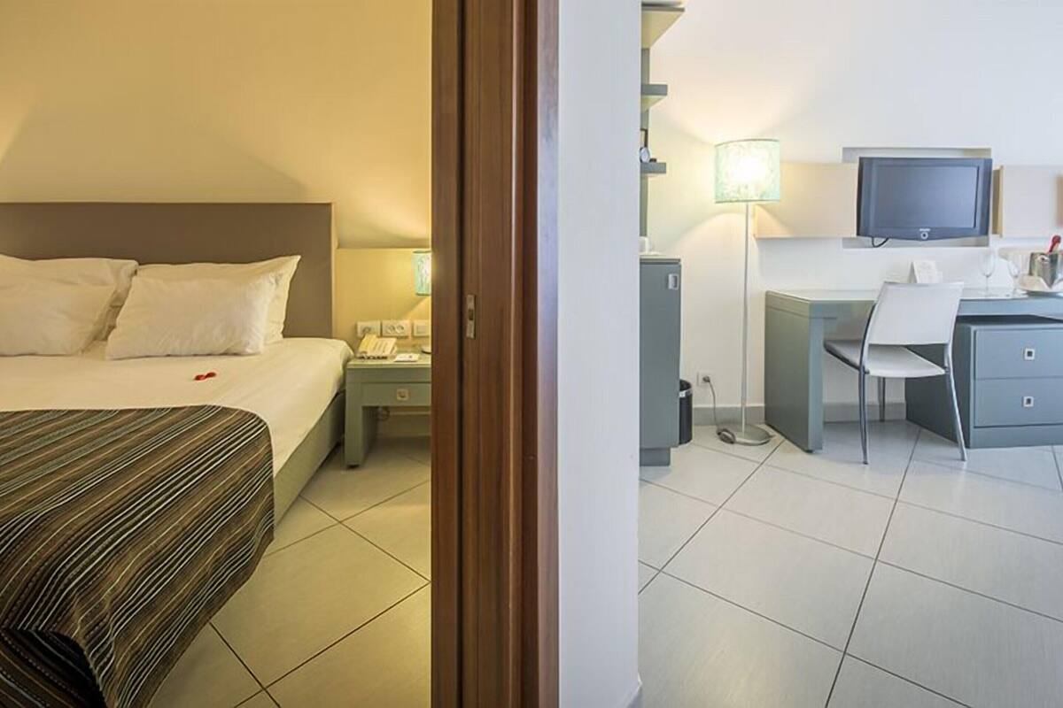 Quarto de hotel com cama, mesas de cabeceira, escrivaninha, cadeira, televisão e abajur.