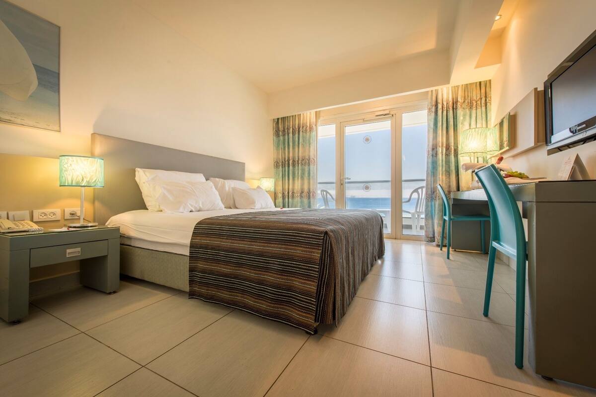 Quarto de hotel com cama, escrivaninha, cadeira, televisão e varanda com cortinas.