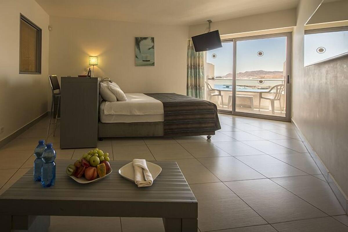 Quarto de hotel com cama, escrivaninha com luminária, mesa com frutas e garrafas, e varanda com vista para a piscina.