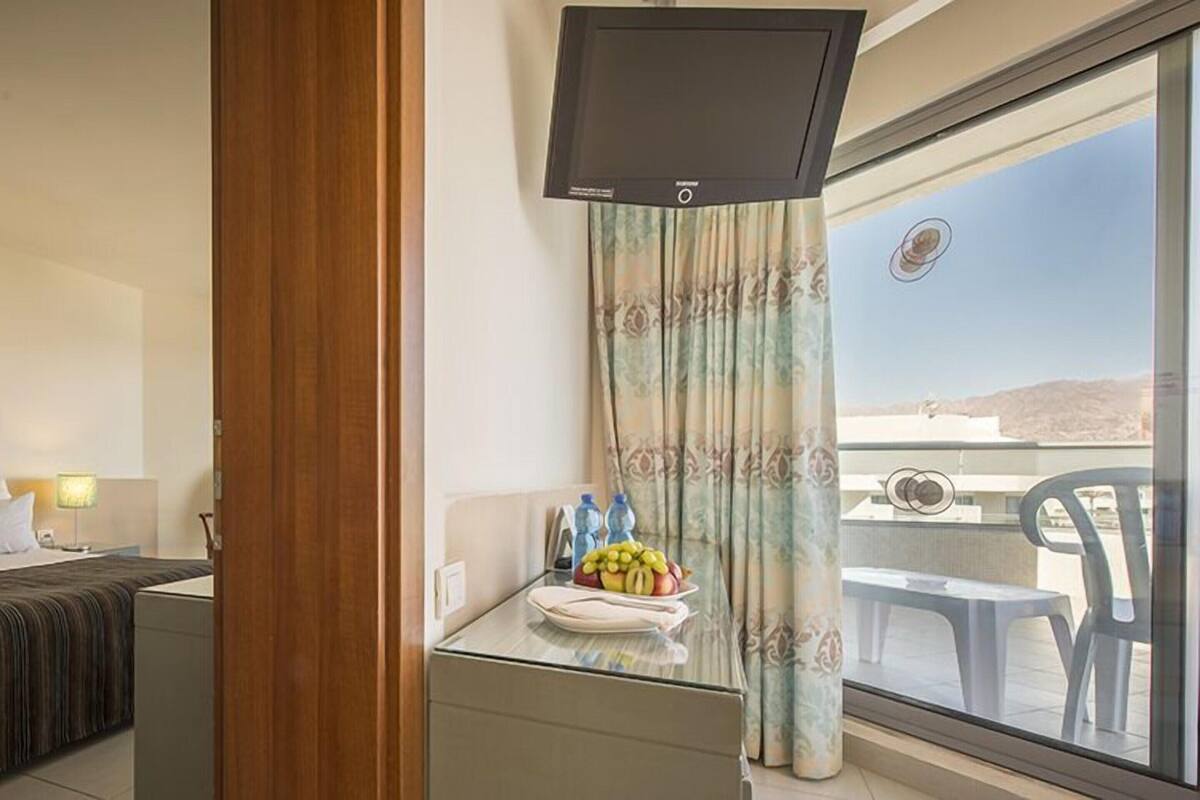 Quarto de hotel com cama, televisão, uma pequena mesa com frutas e garrafas de água, e varanda com vista.