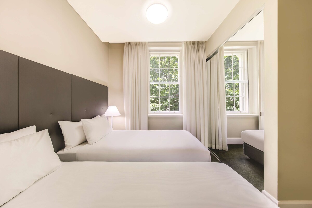 Quarto de hotel com duas camas, uma janela grande com cortinas, uma luminária no teto e cabeceira escura.