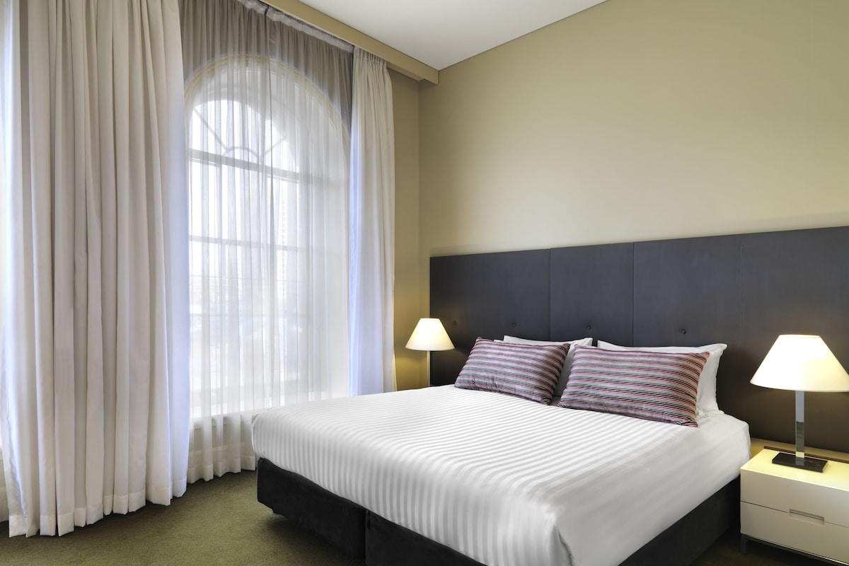 Quarto de hotel com uma cama grande, duas luminárias de cabeceira, uma cabeceira escura e cortinas translúcidas sobre uma janela.