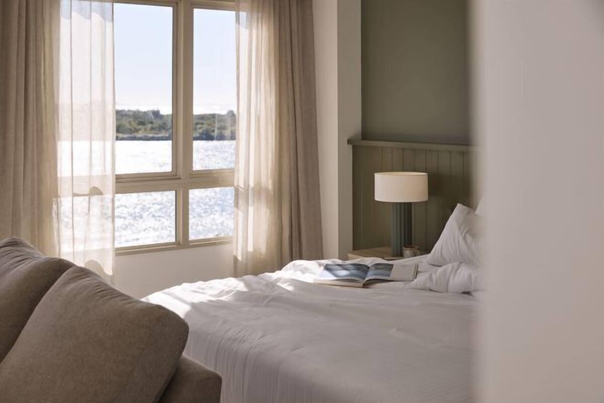 Quarto de hotel com uma cama, mesa de cabeceira, luminária e vista para um corpo de água através de uma janela.