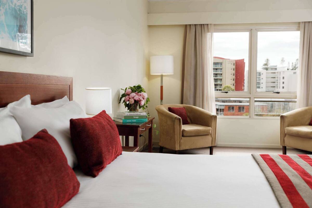 Quarto de hotel com uma cama grande, duas poltronas, uma mesa de cabeceira com abajur, um vaso de flores e vista para prédios pela janela.