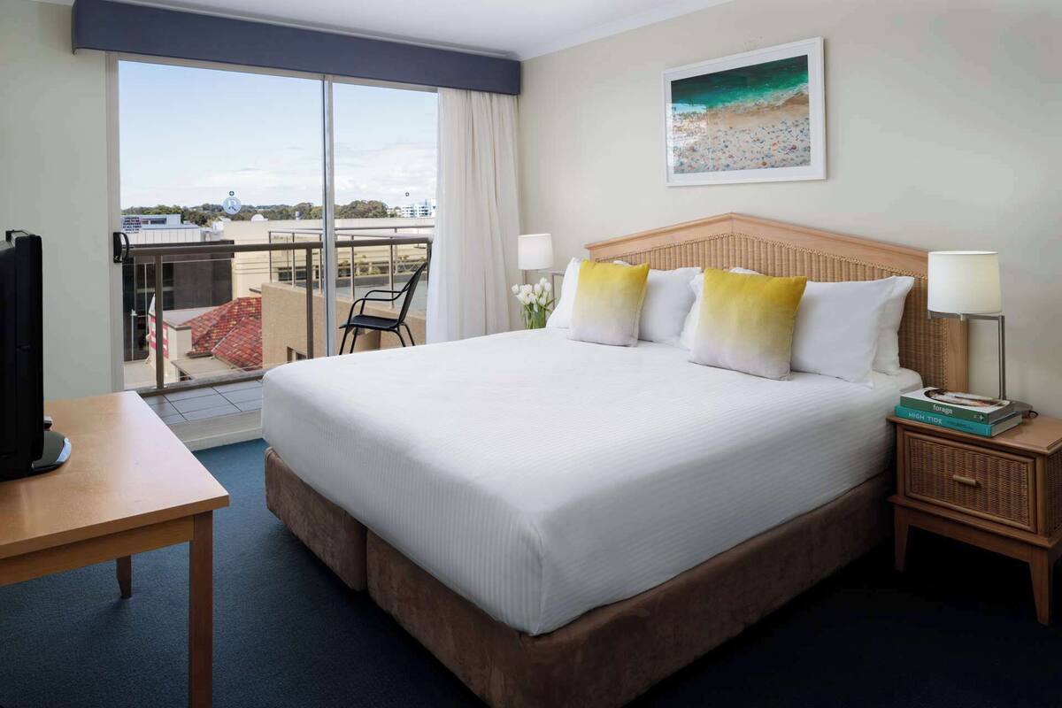 Quarto de hotel com uma cama grande, televisão, escrivaninha, cadeira e varanda com vista para a cidade.