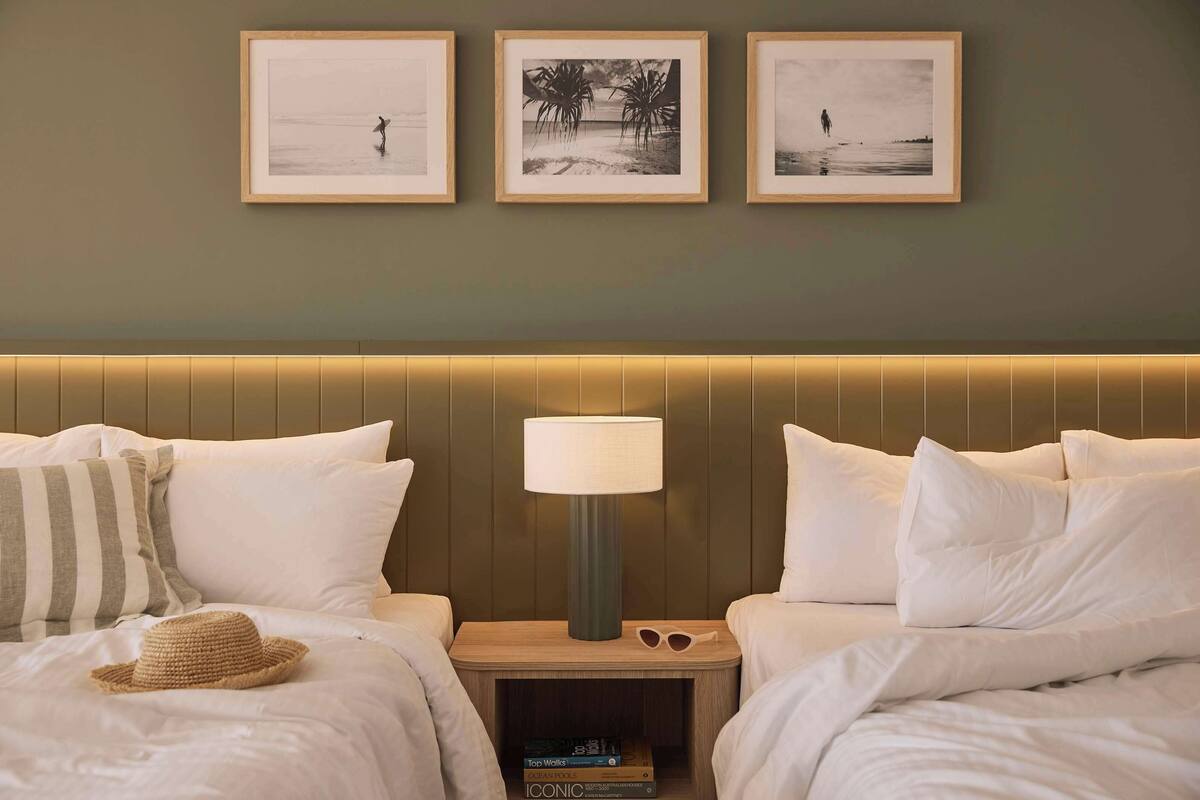 Quarto de hotel com duas camas, uma mesa de cabeceira com abajur e três quadros na parede.