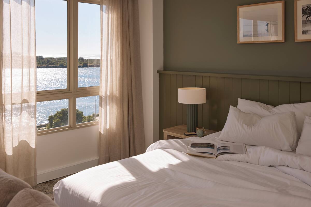 Quarto com cama, mesa de cabeceira, abajur e vista para a água pela janela.