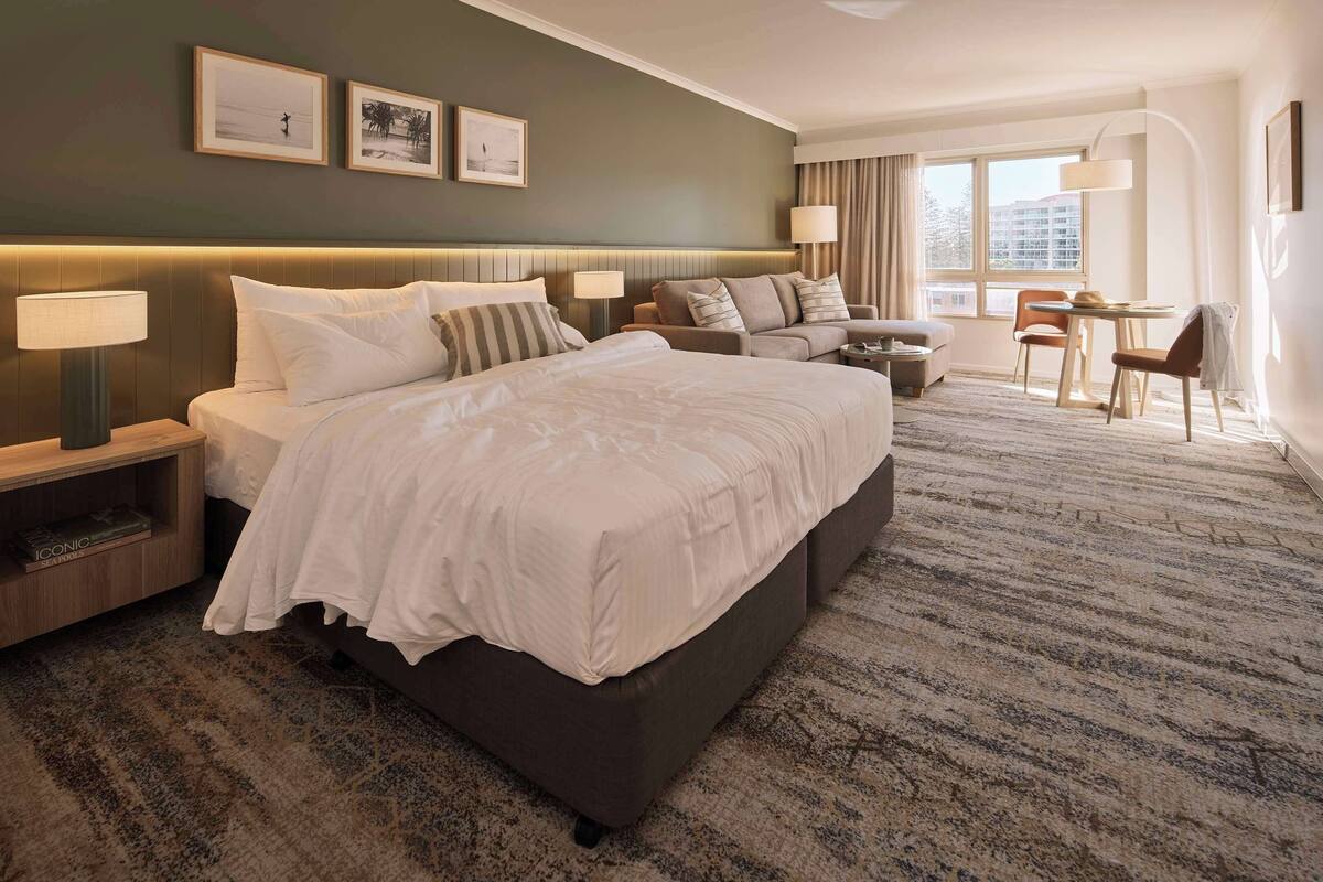 Quarto de hotel com uma cama grande, um sofá, uma escrivaninha com cadeiras e vista para edifícios pela janela.