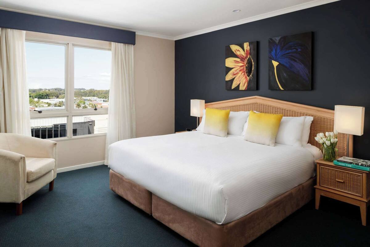 Quarto de hotel com uma cama grande, uma cadeira, mesa de cabeceira com abajur e arte na parede.