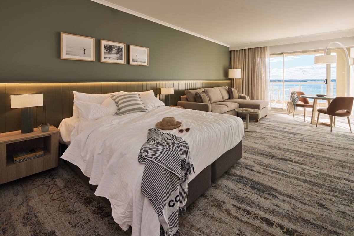 Quarto de hotel com uma cama grande, um sofá, uma mesinha e vista para o mar.