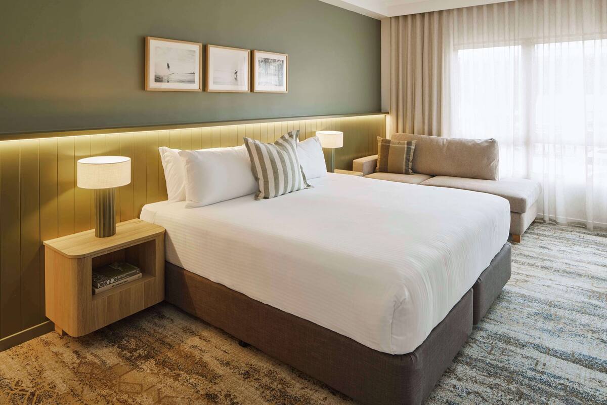 Quarto de hotel com uma cama grande, duas mesas de cabeceira, uma luminária para leitura, um sofá e uma janela com cortinas.