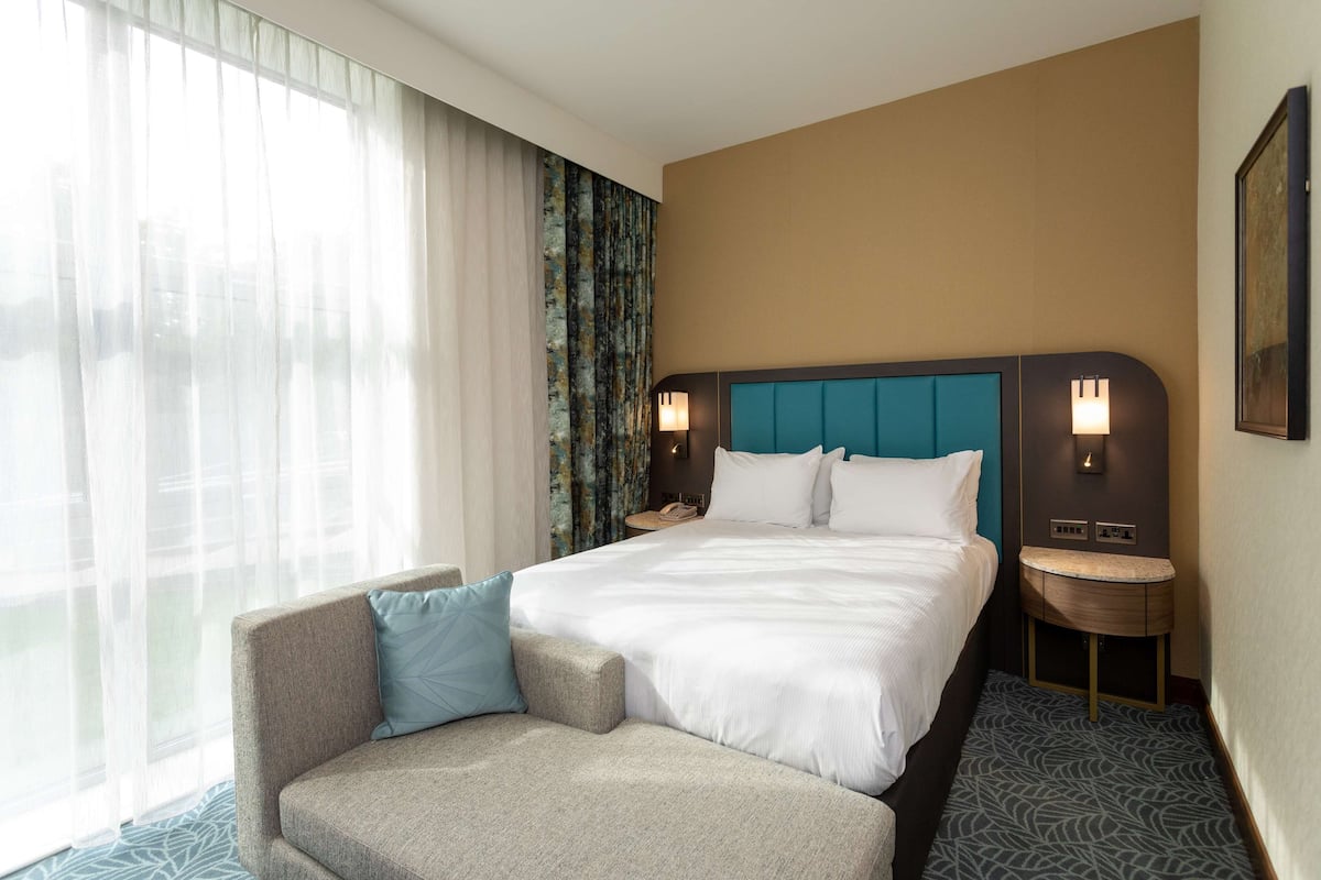 Quarto de hotel com uma cama grande, um sofá, uma mesa de cabeceira com abajur e vista para a cidade através de cortinas finas.