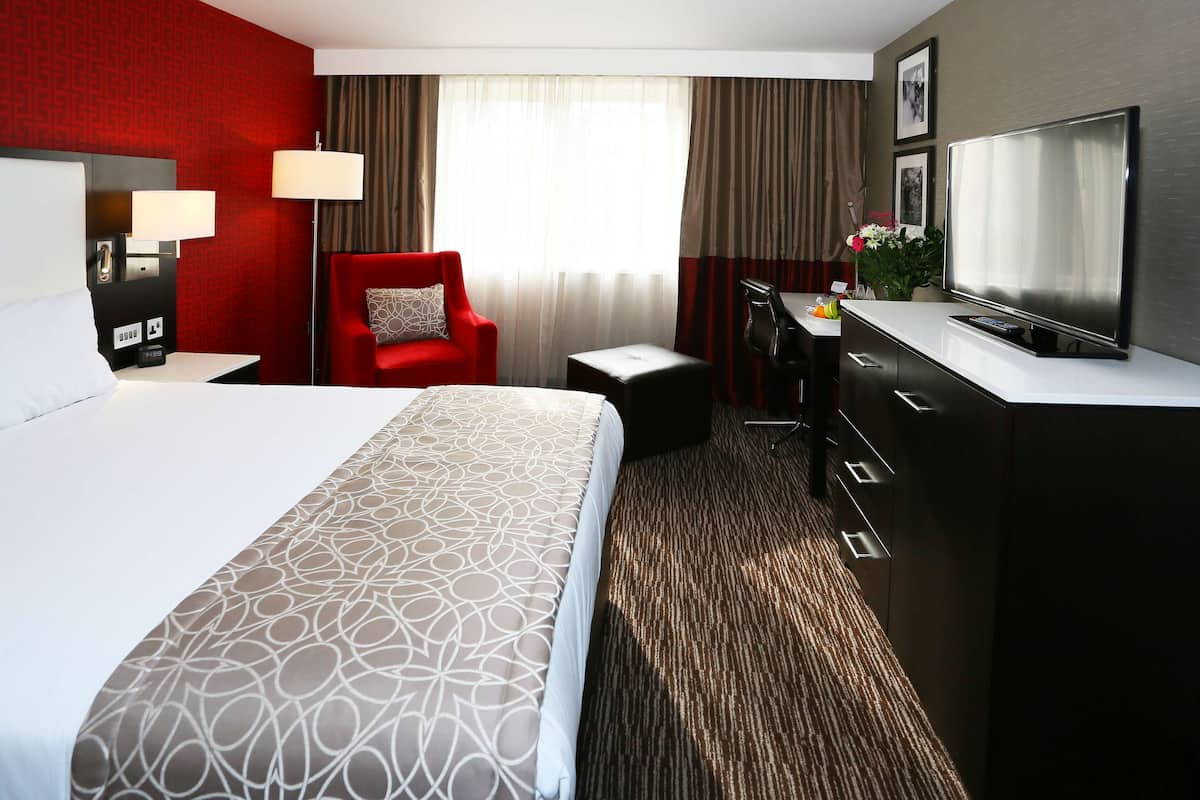 Quarto de hotel com uma cama grande, uma escrivaninha, uma poltrona vermelha, uma televisão e piso coberto por um tapete vermelho.