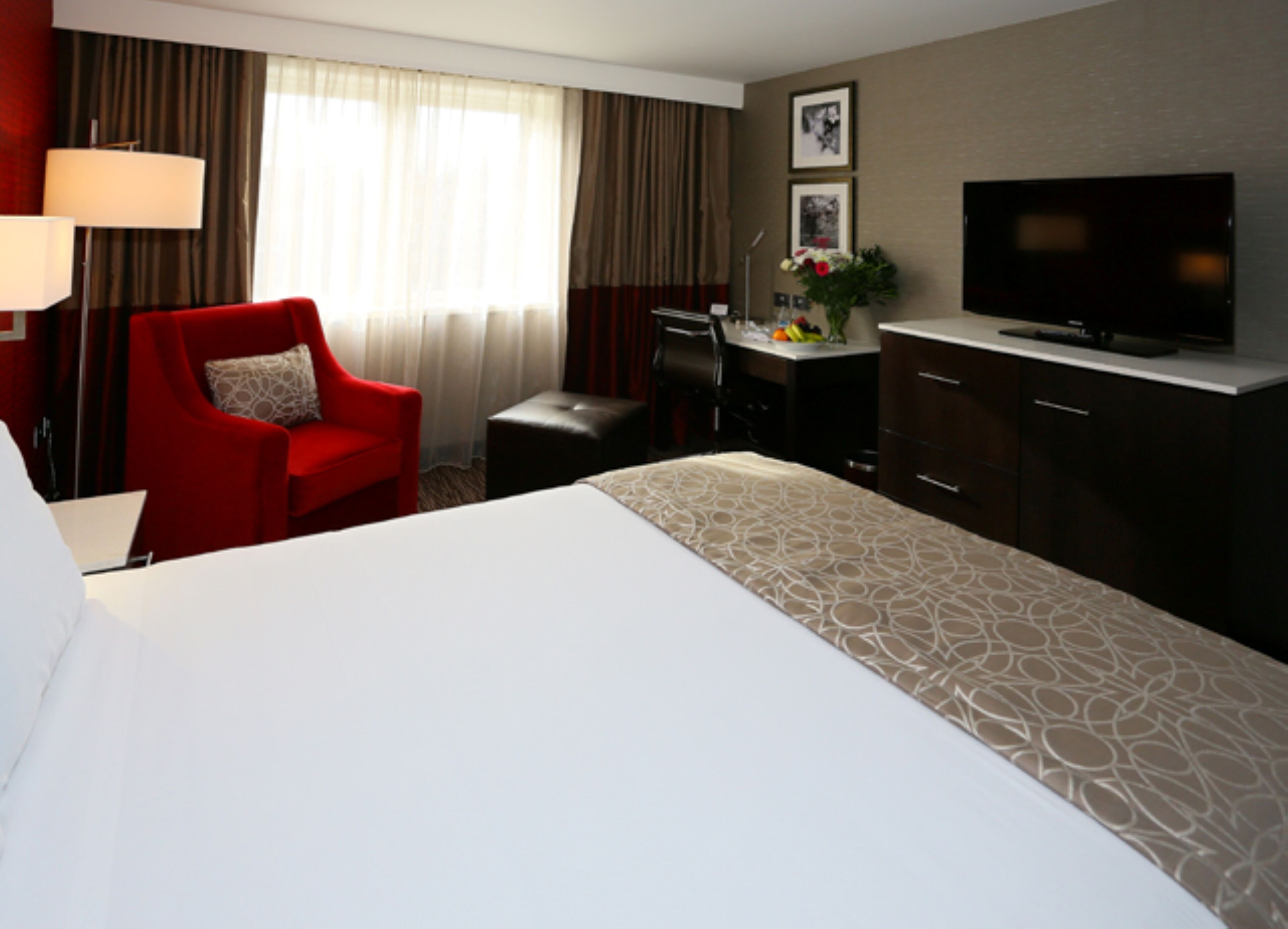 Quarto de hotel com uma cama grande, uma poltrona vermelha, uma escrivaninha com televisão de tela plana e uma janela com cortinas.