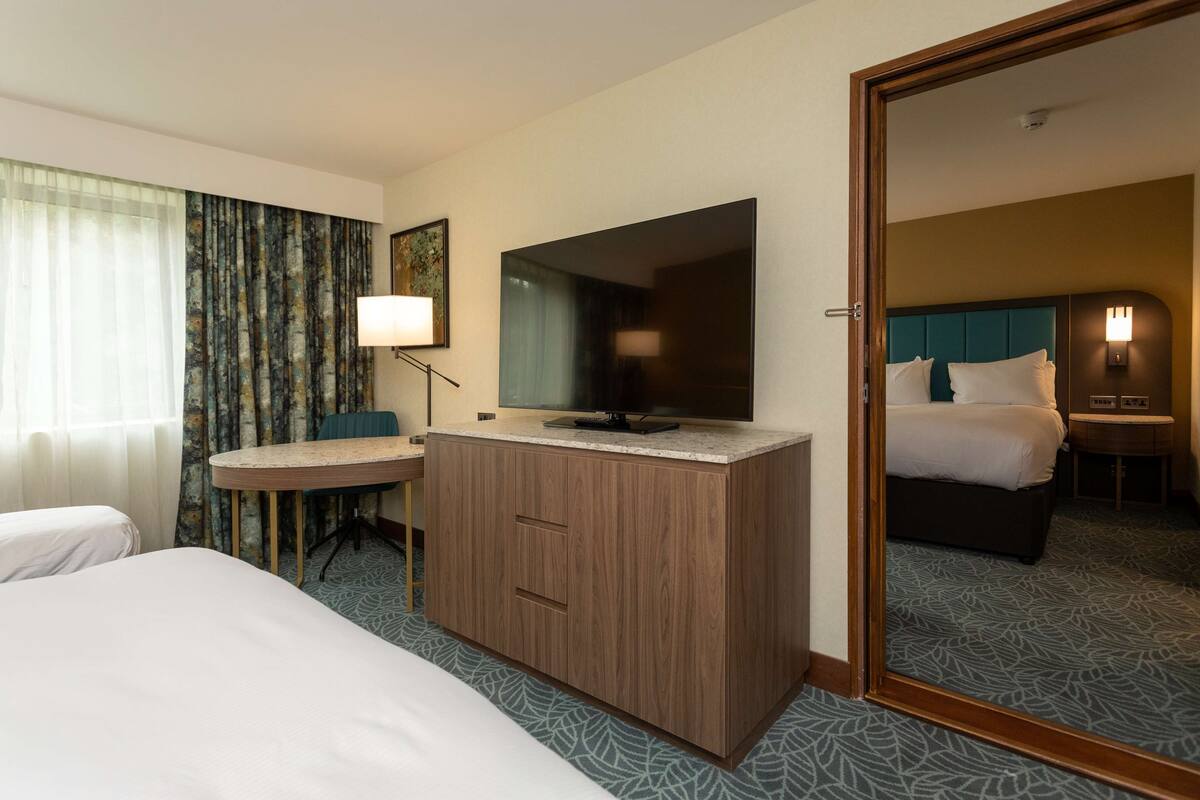 Quarto de hotel com cama, escrivaninha, televisão, espelho e janela com cortinas.