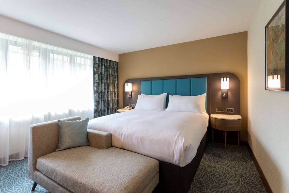 Quarto de hotel com uma cama grande, um sofá, duas mesas de cabeceira com abajures, uma janela com cortinas e um tapete estampado.