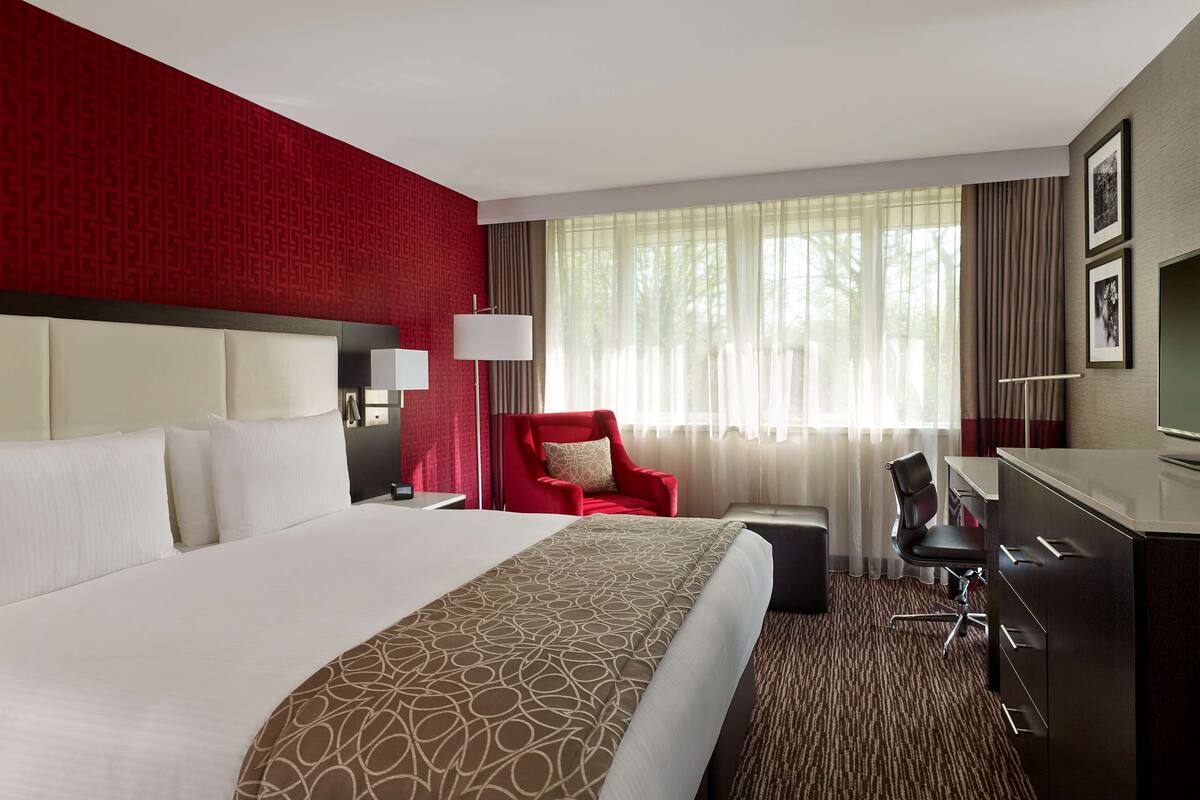 Quarto de hotel com uma cama grande, uma poltrona vermelha, uma escrivaninha e uma janela com cortinas.