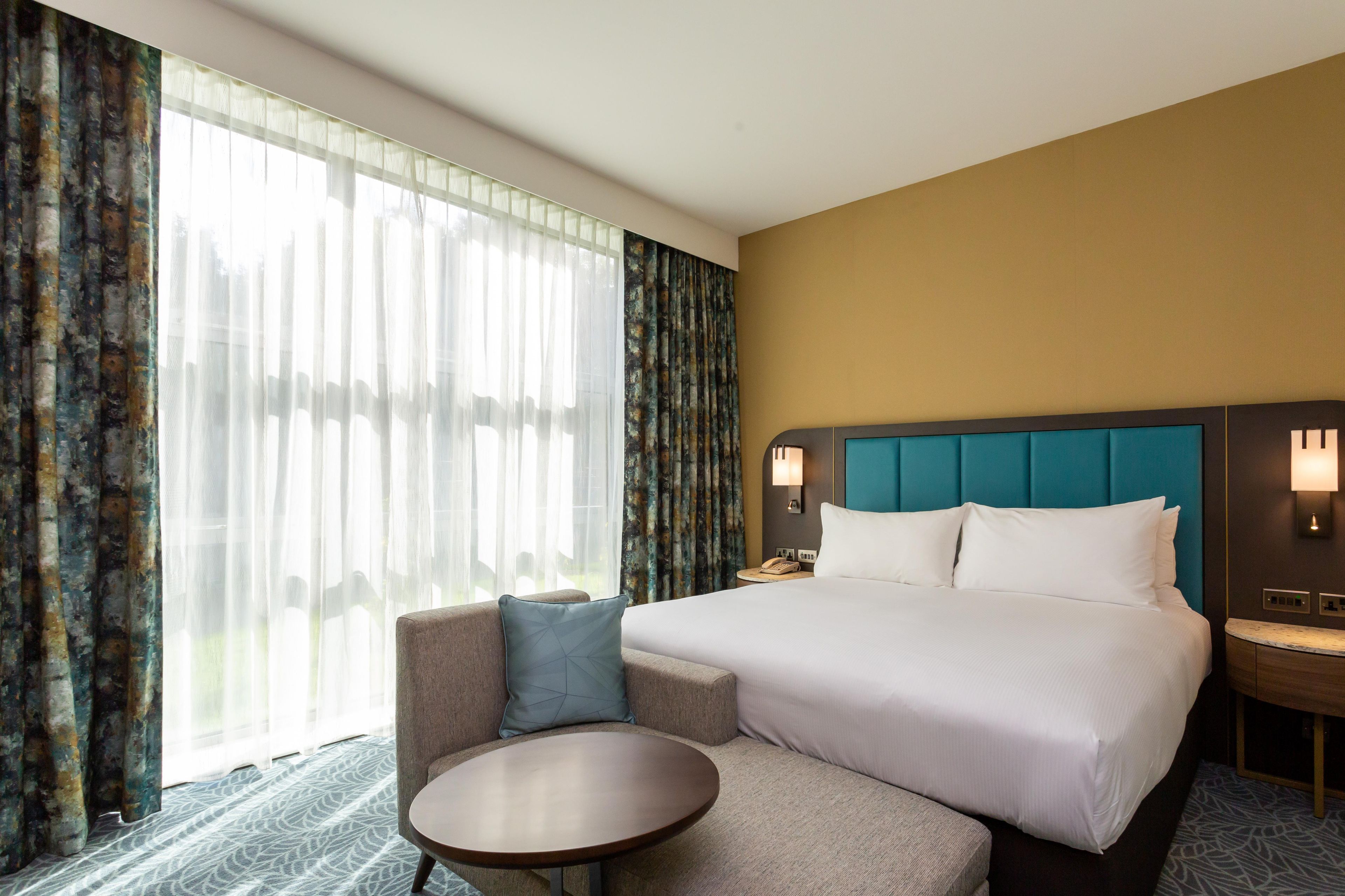 Quarto de hotel com uma cama grande, uma mesa redonda pequena, um sofá e vista para o exterior através de cortinas leves.