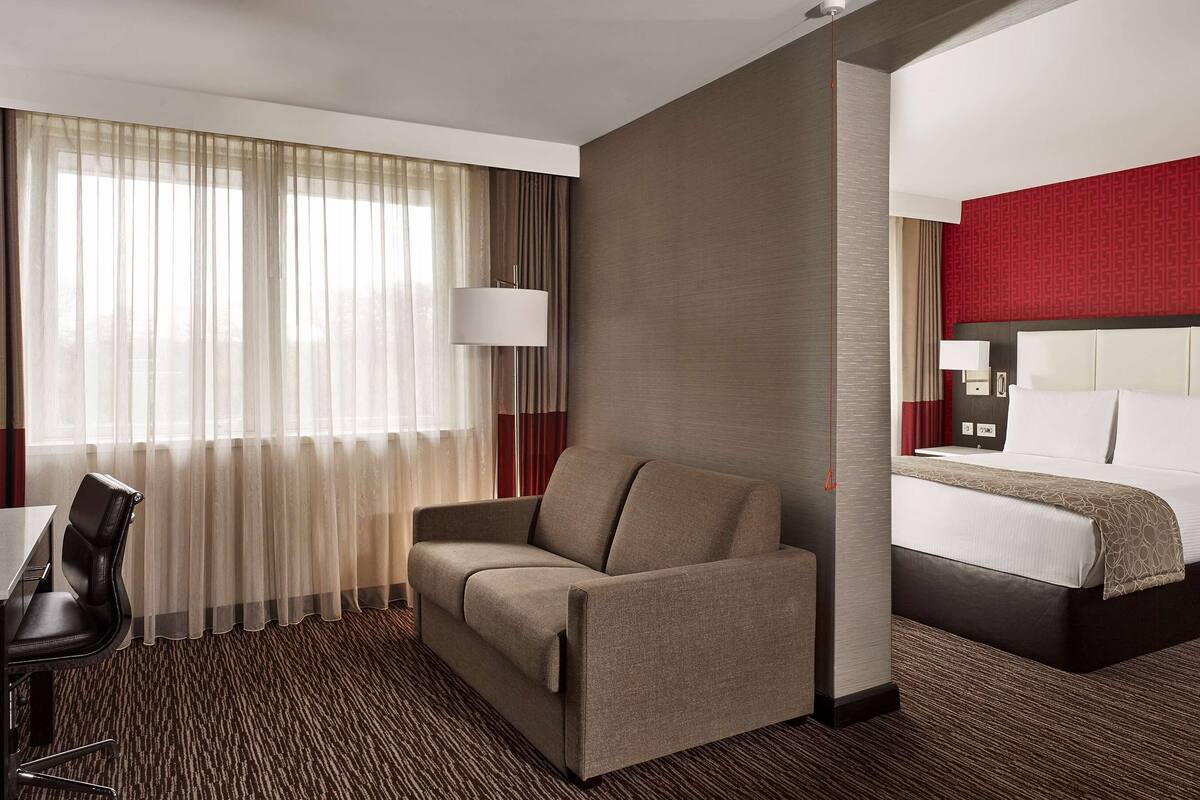 Quarto de hotel com escrivaninha, sofá, cama com parede de destaque vermelha e grande janela com cortinas translúcidas.