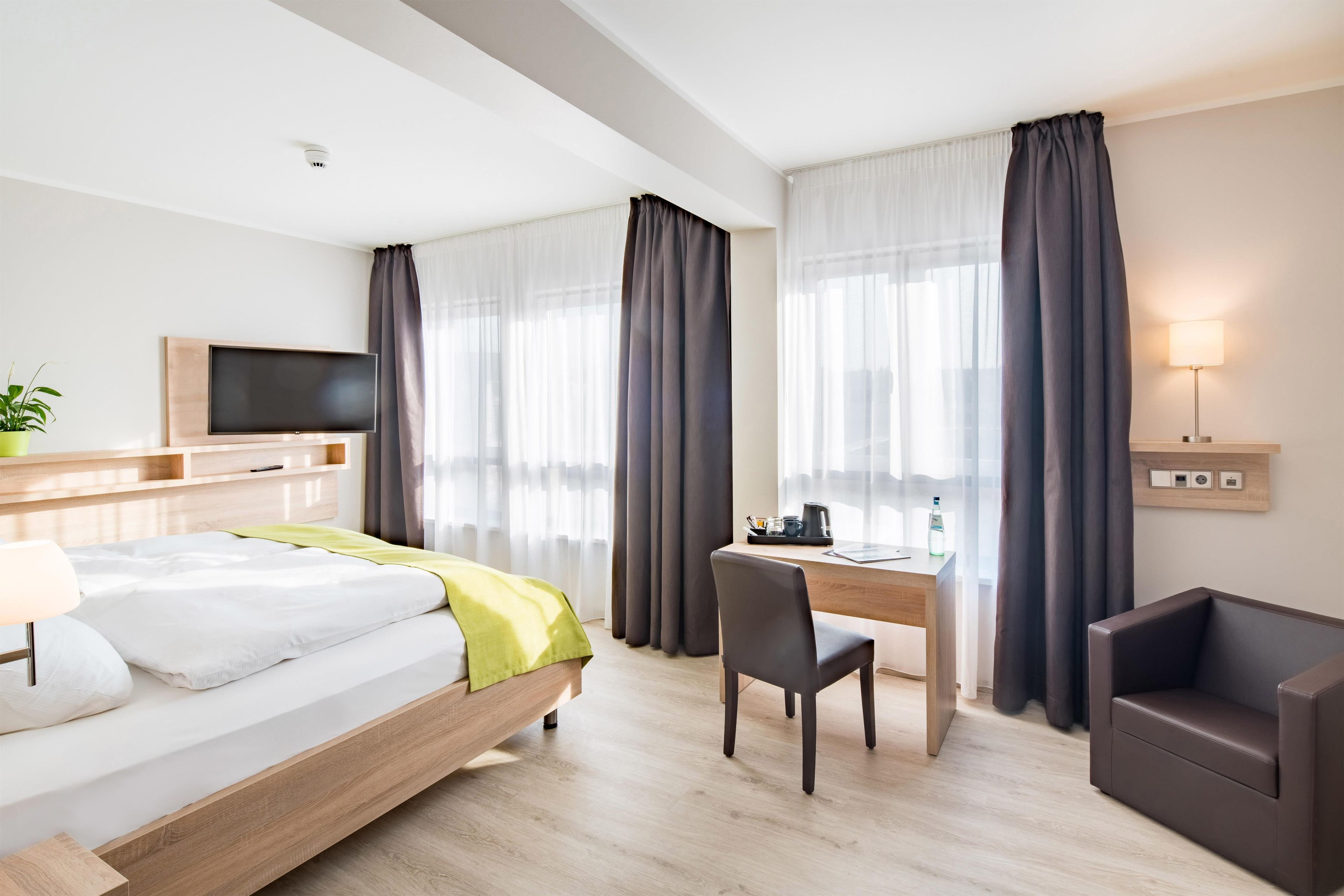 Quarto de hotel com cama, escrivaninha, cadeira e televisão.