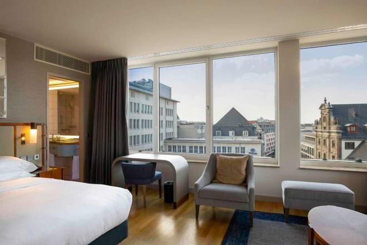 Quarto de hotel com cama, escrivaninha, cadeira e vista para edifícios da cidade.