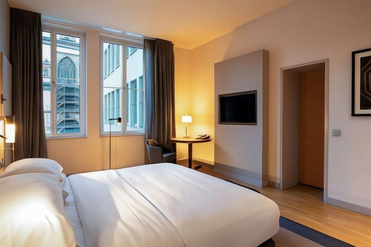 Quarto de hotel com uma cama, escrivaninha, televisão e janela com cortinas.
