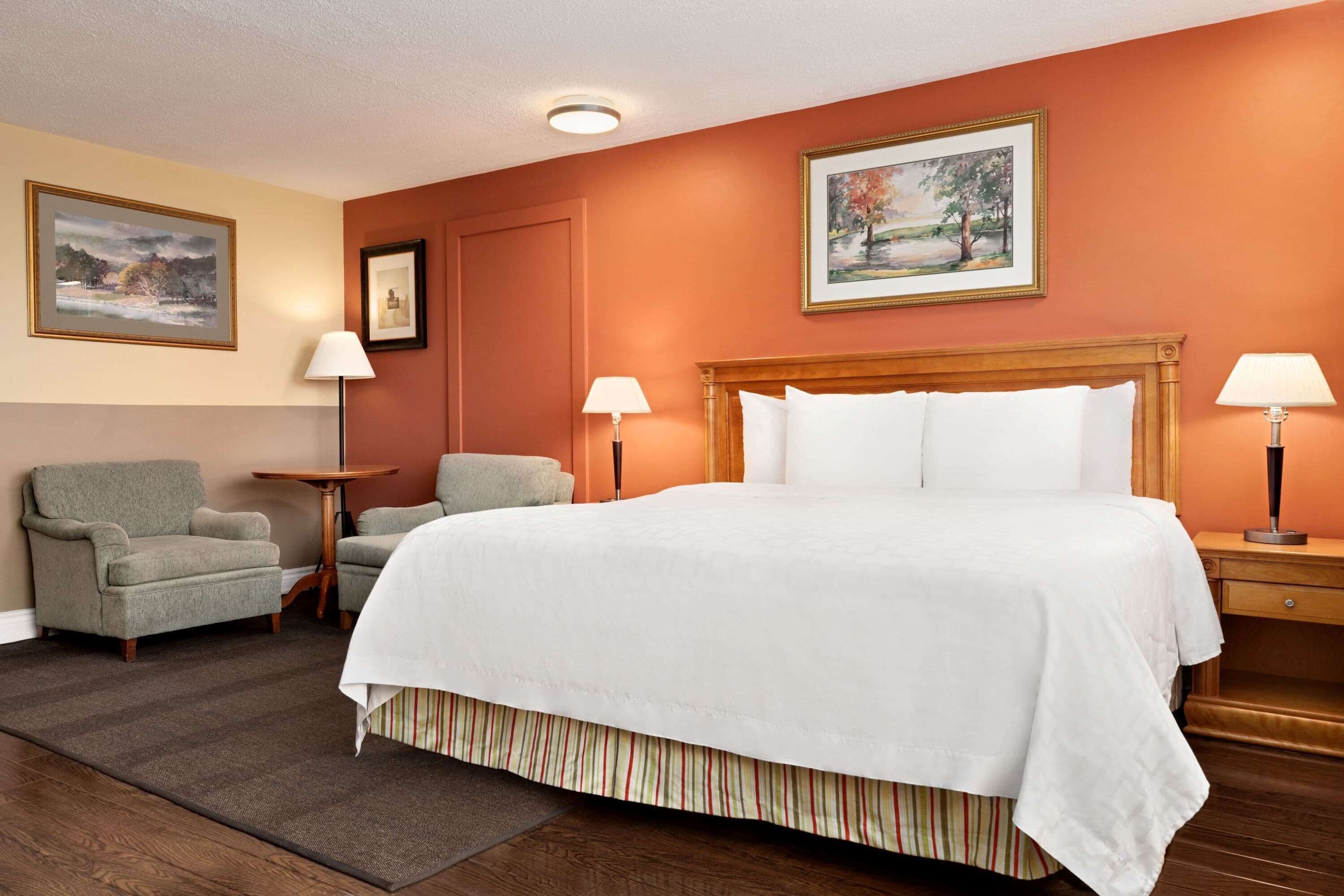 Quarto de hotel com uma cama grande, duas poltronas, uma mesa pequena e uma mesinha de cabeceira com luminárias.
