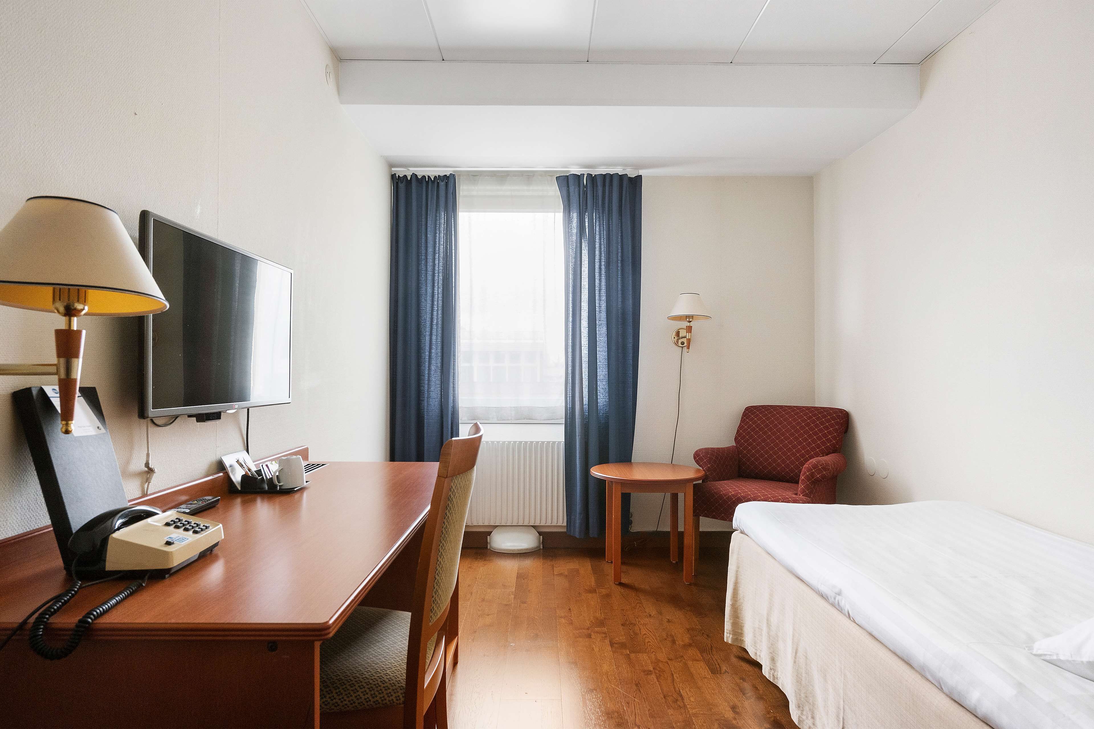 Quarto de hotel com uma escrivaninha de madeira, telefone, televisão, cama, cadeira e uma mesa pequena.