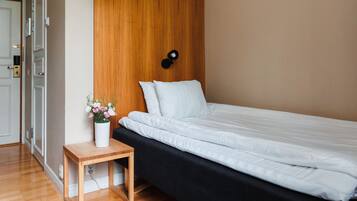 Quarto com cabeceira de madeira, cama branca, mesa de cabeceira pequena de madeira e um vaso de flores, e uma porta à esquerda.