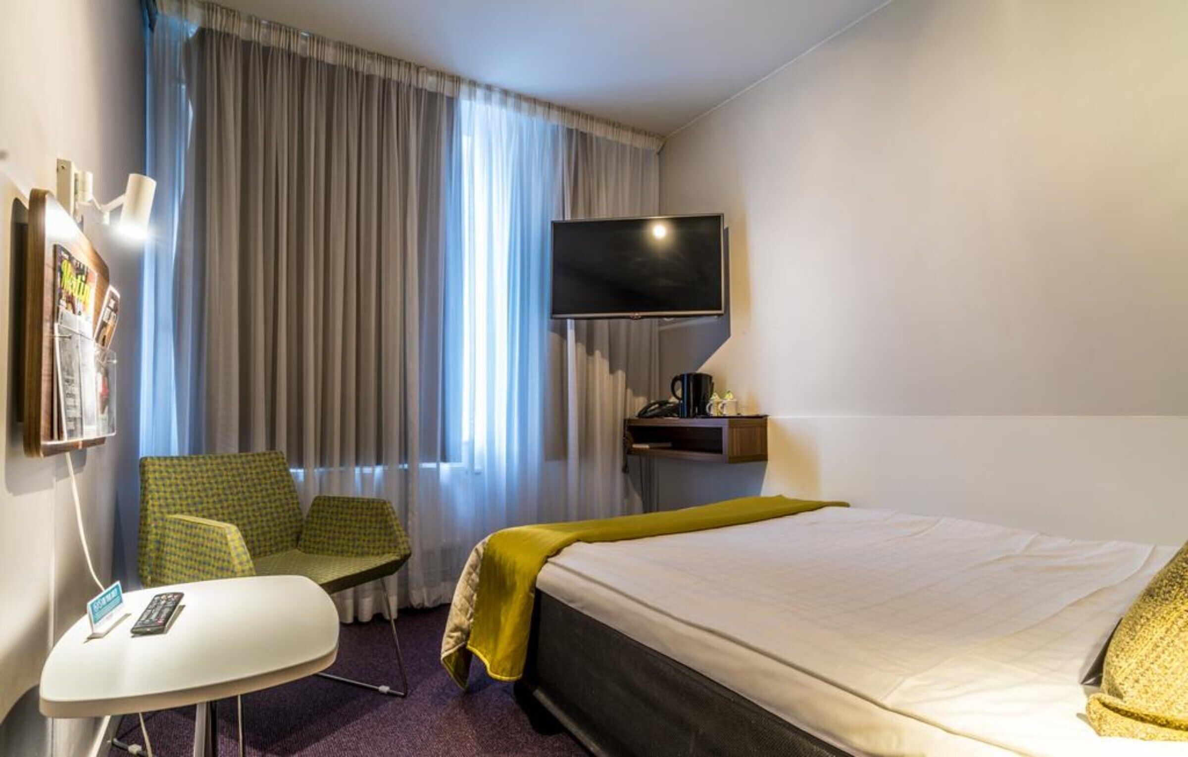 Quarto de hotel com cama, televisão na parede, mesa pequena com controle remoto, cadeira e janela com cortinas.