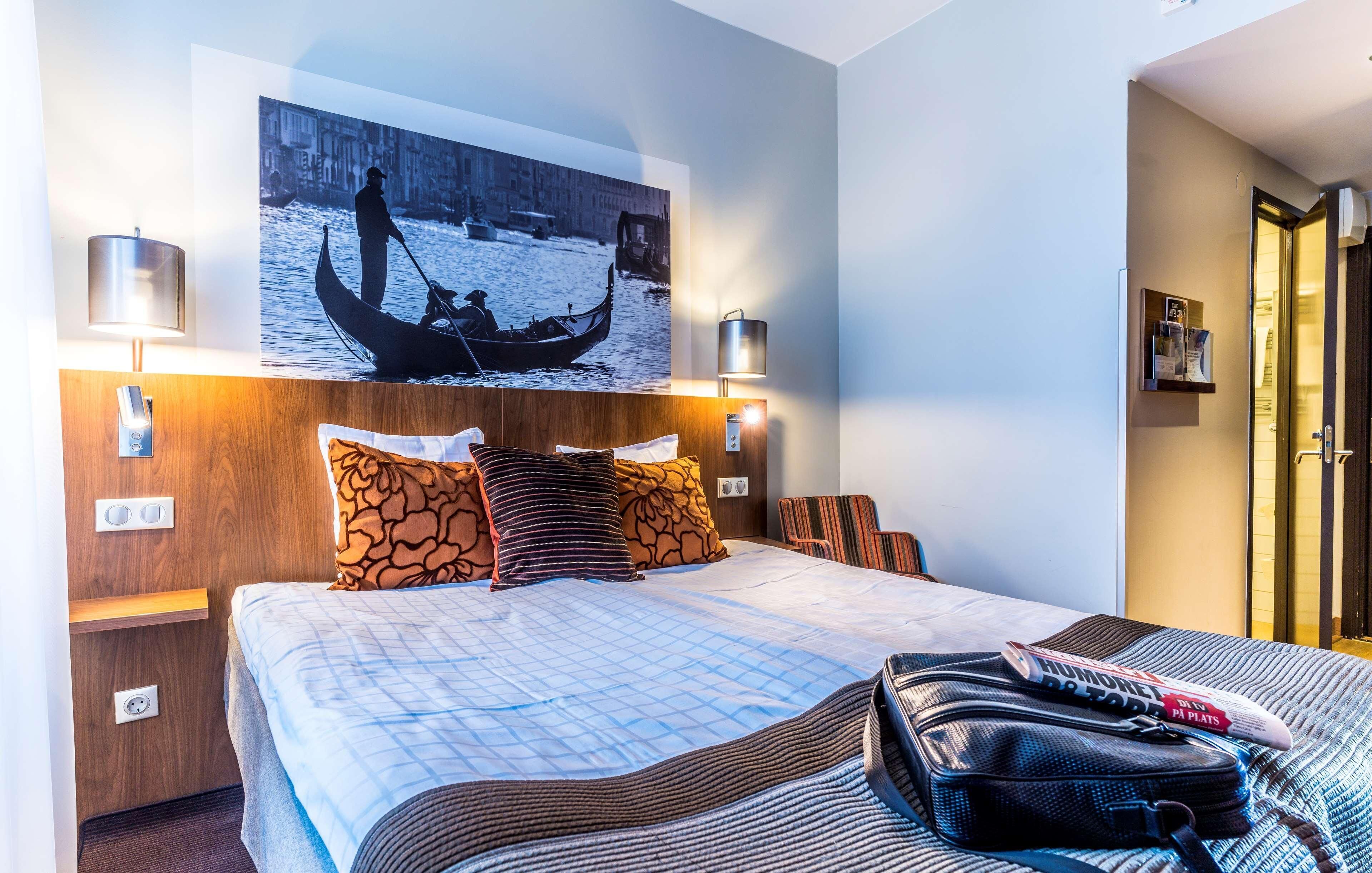Quarto de hotel com cama, cabeceira de madeira, pintura de uma gôndola, duas luminárias de cabeceira, mesa de cabeceira e mala na cama.