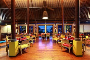 Um restaurante espaçoso com piso de madeira, tetos altos e grandes janelas. As mesas estão dispostas com guardanapos vermelhos e toalhas de mesa brancas.
