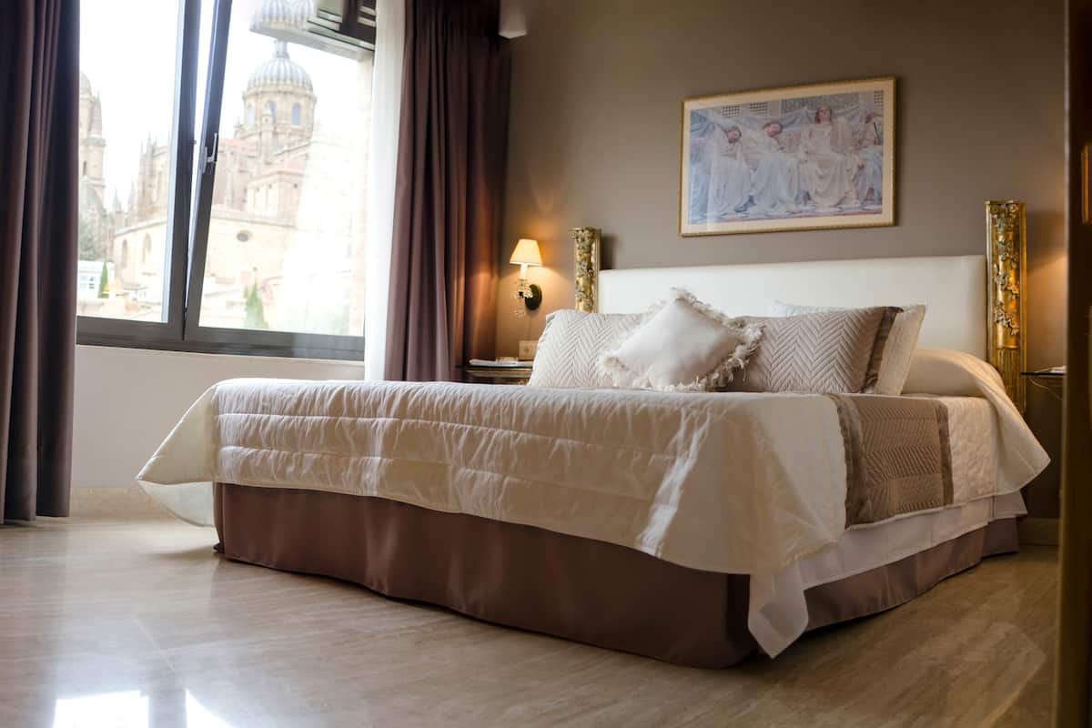 Quarto de hotel com cama, uma cadeira, janela com cortinas, abajur e um quadro na parede.