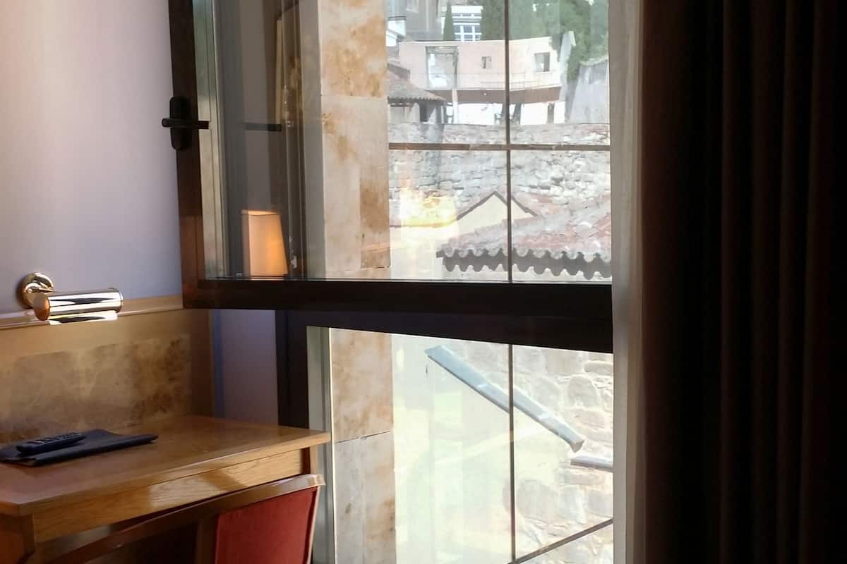 Quarto com escrivaninha de madeira, uma cadeira vermelha e uma janela ampla com vista para um templo.