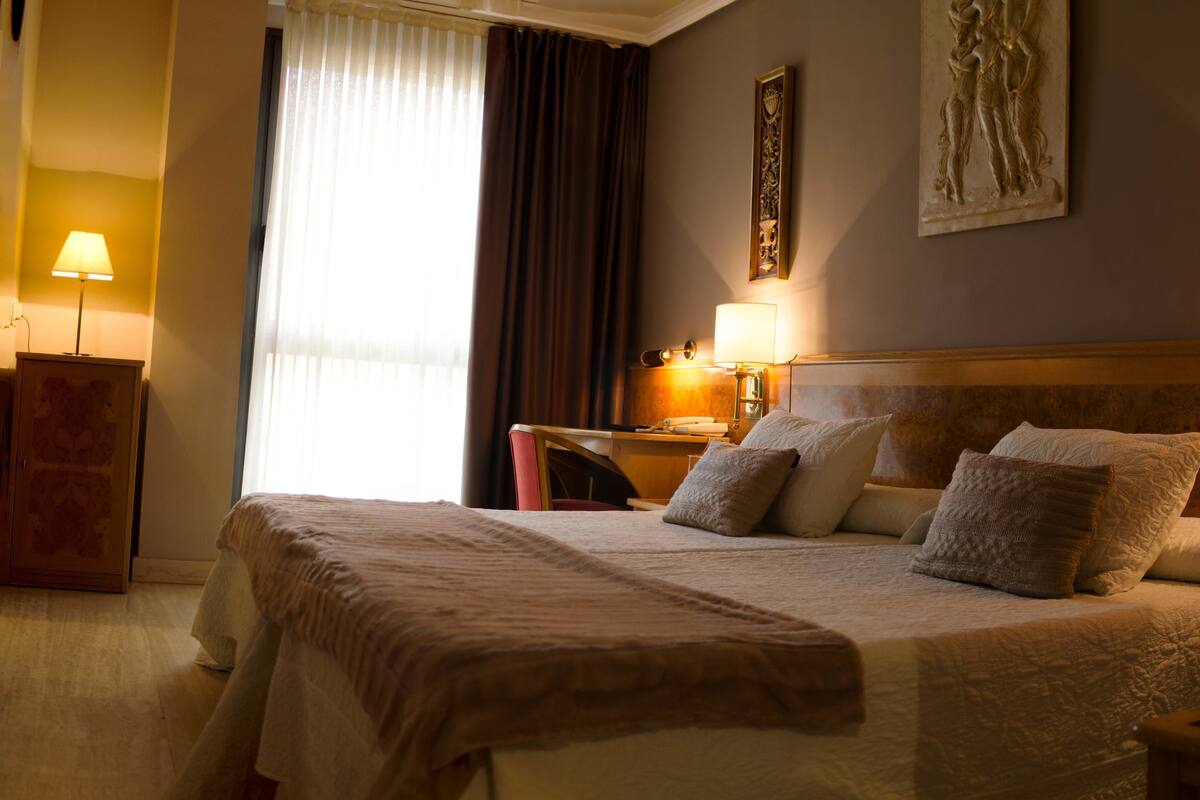 Quarto de hotel com uma cama grande, mesa de cabeceira, luminária, escrivaninha e janela com cortinas.