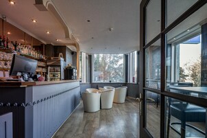 Uma área de bar com balcão de madeira, prateleiras com garrafas e uma janela ampla com vista para árvores.
