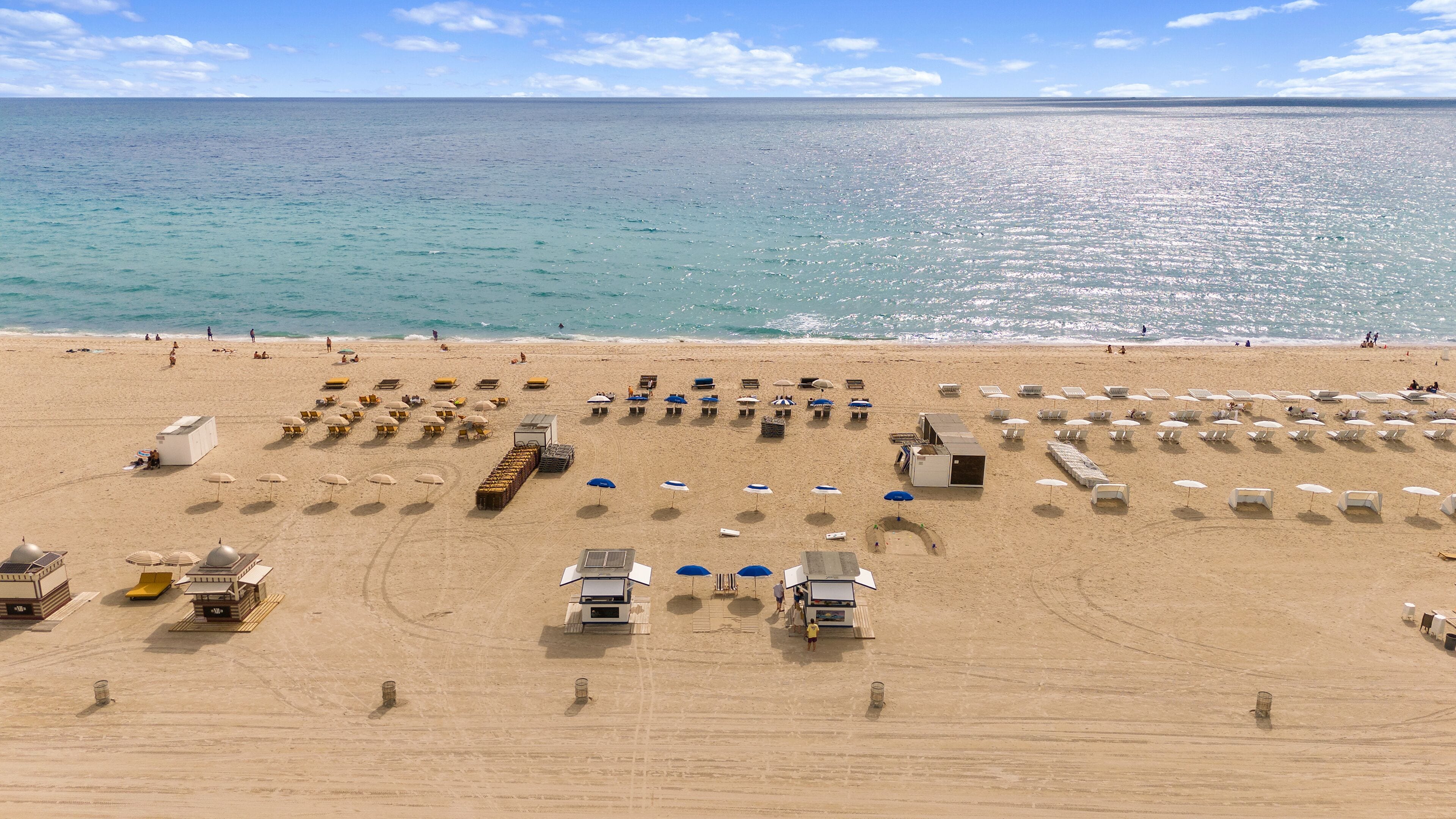 Uma praia com fileiras de espreguiçadeiras e guarda-sóis, algumas cabanas de praia e uma vista ampla do oceano.