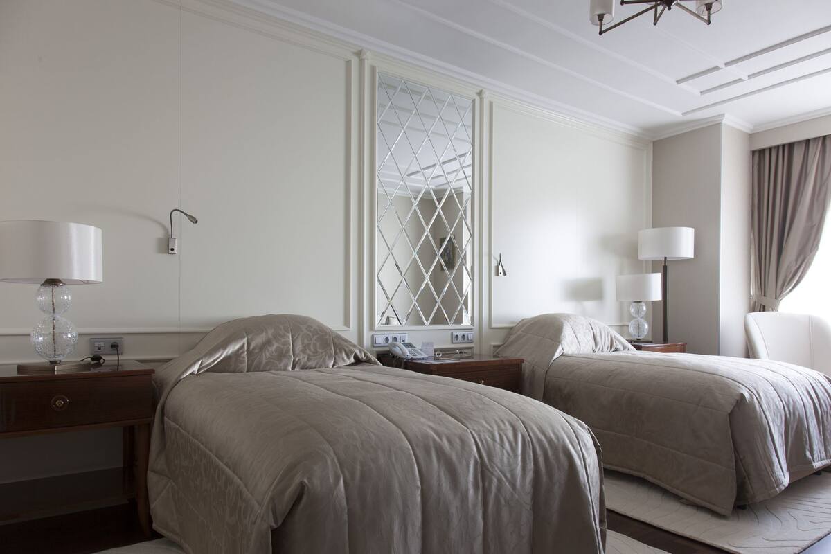 Quarto de hotel com duas camas, uma janela grande com padrão de losangos, luminárias de cabeceira e um telefone na mesinha de noite.