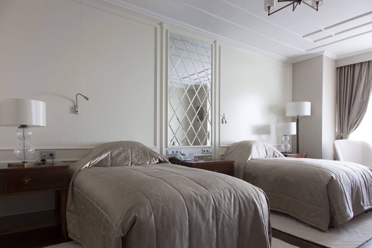 Quarto de hotel com duas camas, uma janela com grade em padrão de losangos, luminárias de cabeceira, um telefone e uma mesa de cabeceira.