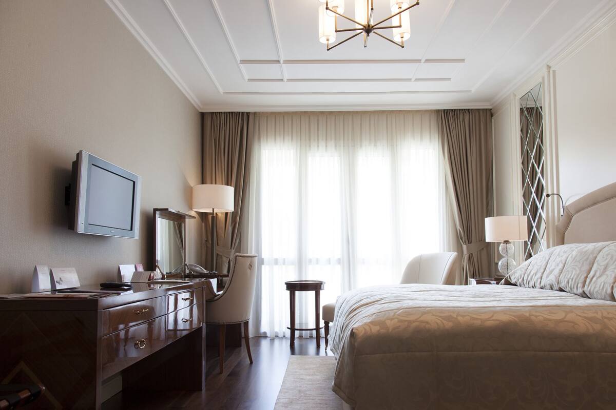 Quarto de hotel com cama, escrivaninha, cadeira, televisão e janela com cortinas.