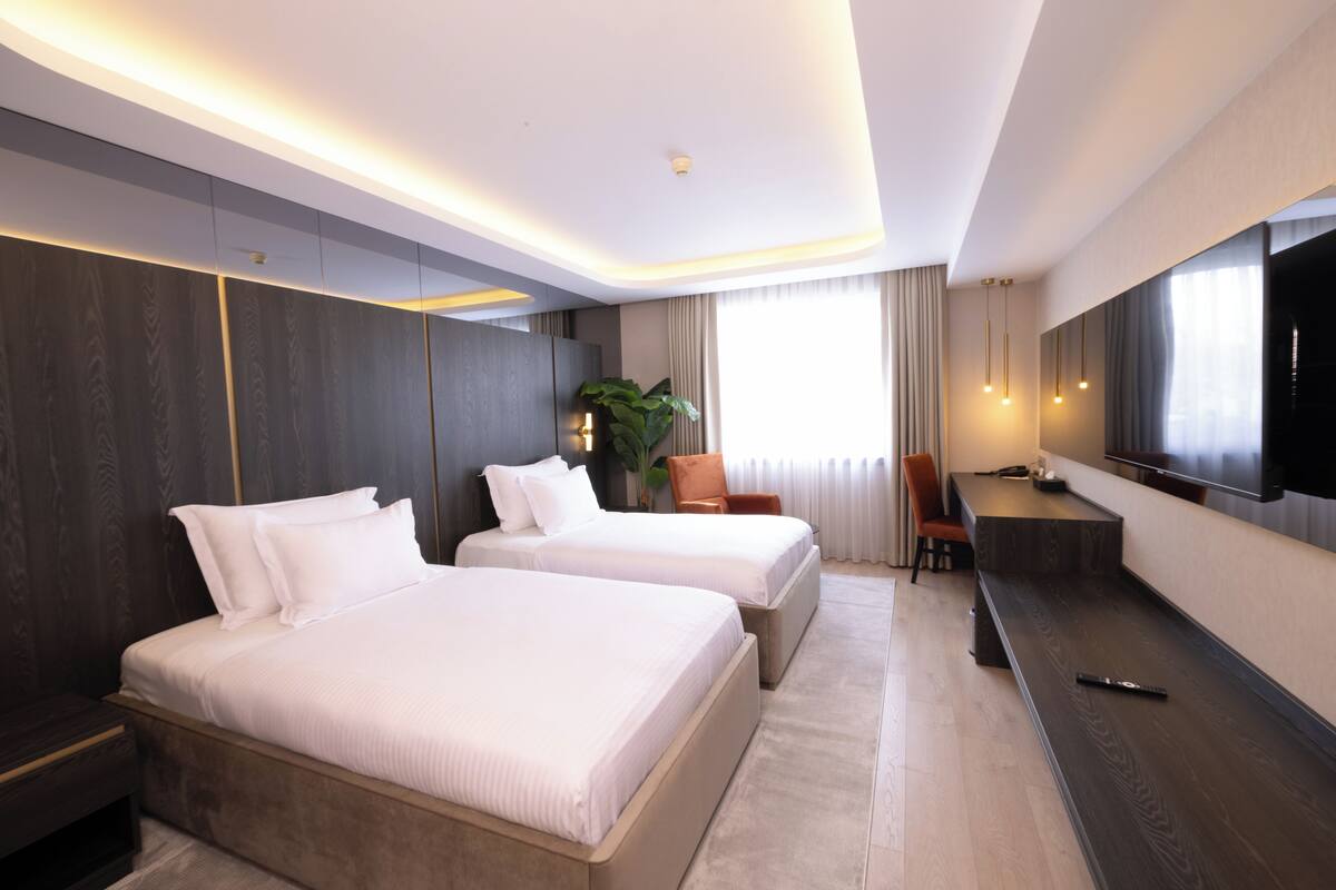 Quarto de hotel moderno com duas camas, uma escrivaninha, uma cadeira, televisão e uma janela grande.